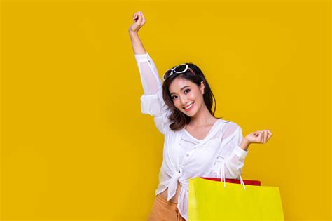 노란 스튜디오 배경에 고립 된 쇼핑 백을 들고 쾌활웃고 웃고 있는 아시아의 아름다운 행복한 젊은 여성 행복 소비주의 판매 및 쇼핑 개념 가방에 대한 스톡 사진 및 기타