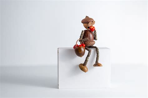 도토리 나뭇가지 씨앗과 같은 천연 재료로 만든 작은 조각상입니다 조각상은 빨간 열매로 가득 찬 작은 바구니를 들고 흰색 상자 위에 앉아 있습니다 Diy에 대한 스톡 사진 및