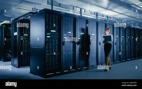 In Data Center Female It Technician Walks Alongside Row Of Server Racks Uses Laptop Computer