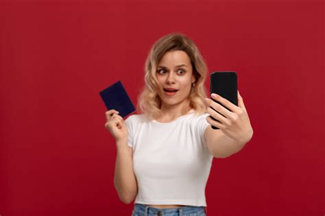 빨간 배경에 흰색 티셔츠에 곱슬 금발 머리를 가진 여자의 초상화입니다 해피 모델은 여권을 들고 휴대 전화로 셀카를 만든다 T 셔츠에 대한 스톡 사진 및 기타 이미지 Istock