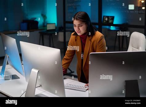 Portrait Of Female Cybersecurity Expert Using Computer With Multiple Monitors In Office Lit By