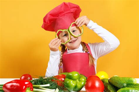 요리사의 모자를 쓴 명랑한 아이는 채소와 함께 재미를 가지고 있으며 마치 안경을 쓴 것처럼 눈 앞에서 후추를 들고 있습니다 노란 벽의 배경에 테이블에 앉아 어린 소녀 눈