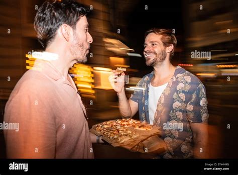 A Joyful Gay Couple Shares A Slice Of Pizza While Walking And Laughing Together In A Bustling