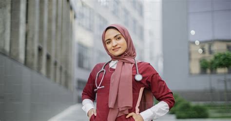 현대 클리닉 밖에서 히잡 스크럽 청진기를 착용한 아름다운 말레이 여의사 야외에 서 있는 빨간 스크럽을 입은 친절하고 웃고 있는 자신감 있는 무슬림 여성 의사의 클로즈업