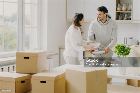 바쁜 가족 부부의 사진은 포장 상자에서 개인 물건을 풀고 캐주얼 한 옷을 입고 흰색 접시를 잡고 패키지 더미로 둘러싸인 현대적인 가구가있는 넓은 주방에서 포즈를 취합니다 2