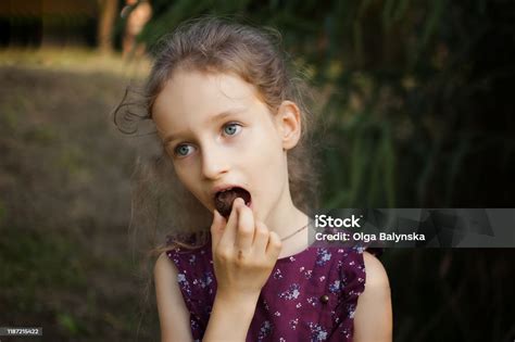 귀여운 작은 금발 소녀 와 포니 테일과 보라색 드레스 먹는 초콜릿 사탕 을 먹는 공원에서 하루 를 괴롭히는 건강에 해로운 라이프 스타일 개념 손가락에 대한 스톡 사진 및