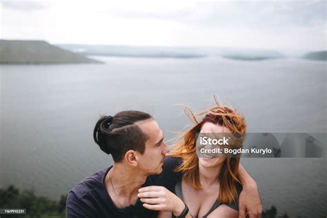 행복한 힙스터 부부는 앉아서 강에 아름다운 전망과 바위 산의 꼭대기에 웃음 바람이 부는 절벽에서 포옹하고 미소 짓는 관광 부부 함께 여행 복사 공간 강에 대한 스톡 사진 및