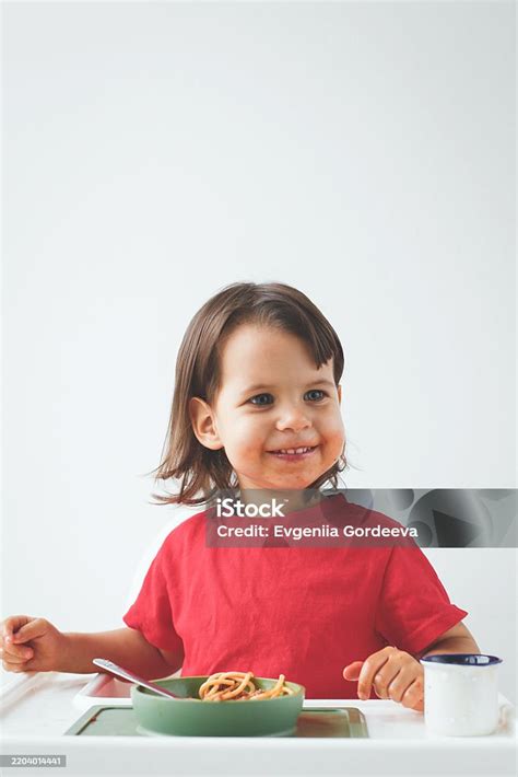 빨간 셔츠를 입은 웃는 아이가 스파게티 한 그릇과 작은 컵을 들고 높은 의자에 앉아 흰색 배경에 남은 음식을 얼굴에 묻힌 채