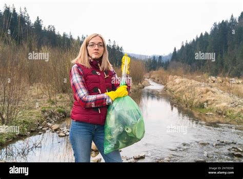Blond Hair Woman In Glasses Collecting Plastic Garbage Near The River And Mountains Forest