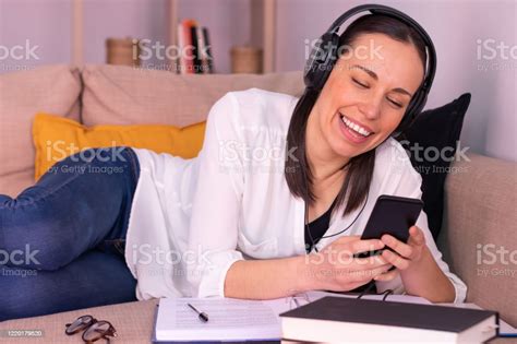 예쁜 갈색 머리 젊은 여자는 헤드폰을 통해 음악을 듣고 집에서 소파에 뻗어 공부 하는 그녀의 스마트 폰으로 웃고 Mp3 플레이어에 대한 스톡 사진 및 기타 이미지 Istock