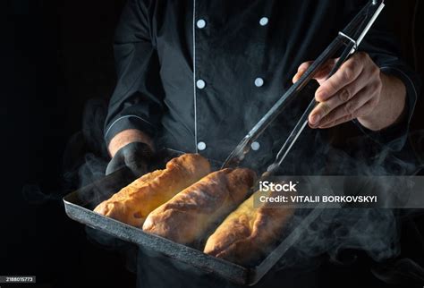 검은 코트를 입은 셰프가 쟁반에서 김이 모락모락 나는 황금빛 롤을 조심스럽게 들어 올려 아늑한 주방 분위기에서 따뜻함과 풍미를 약속하는 맛있는 요리를 선보입니다 가정 주방에