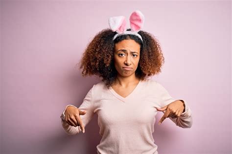 분홍색 배경 위에 토끼 귀를 입은 아프리카 계 미국인 젊은 여성은 슬프고 화가 나고 손가락으로 방향을 가리키고 불행하고 우울합니다 가리키기에 대한 스톡 사진 및 기타 이미지