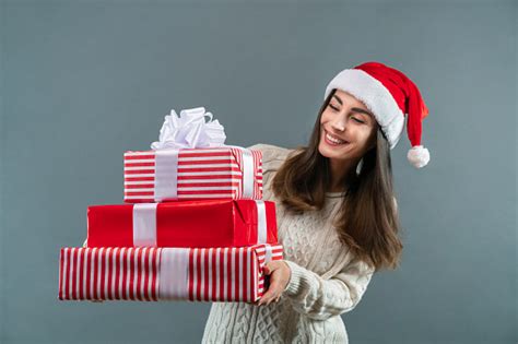 회색 배경 위에 고립 된 손에 장식 된 선물 상자 더미와 함께 포즈를 취하는 산타의 모자에서 행복한 미소 아름다운 소녀의 초상화 25 29세에 대한 스톡 사진 및 기타