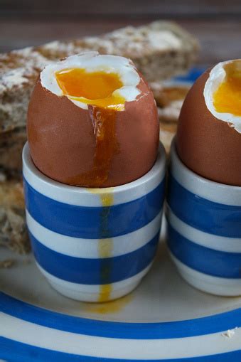 파란색과 흰색 코니쉬 도자기 그릇 차 접시 달걀 컵 부드러운 삶은 달걀 통밀 버터 구운 빵 조각 콧물 노란색 노른자 나무 곡물 배경 전경에 초점을 맞춘 계란을 얹은 풀