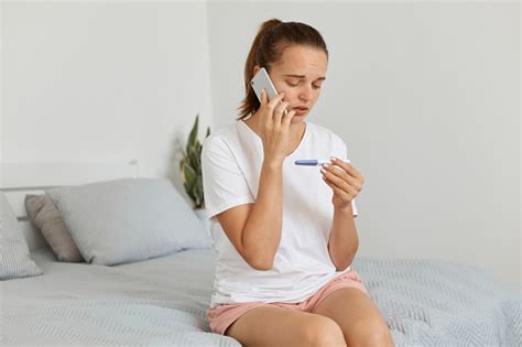 임신 검사를 보고 얼굴에 화가 난 표정으로 침대에 앉아 있는 슬픈 여성의 실내 사진은 흰색 티셔츠와 짧은 티셔츠를 입고 그 결과에 대한 슬픔을 느낍니다 전화에 대한 스톡