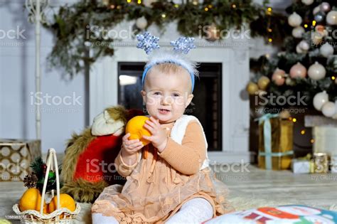 크리스마스 벽난로와 크리스마스 트리 근처 푹신한 카펫에 앉아 쾌활한 소녀 그녀는 손에 귤을 들고 카메라를 바라본다 가로 방향입니다 크리스마스의 개념 12월에 대한 스톡 사진