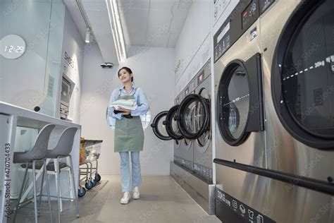 셀프 서비스 세탁실에서 건조기와 세탁기를 배경으로 빨래를 들고 서있는 젊은 아시아 한국 여자 모델 фотография Stock Adobe Stock
