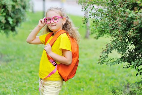 노란색 티셔츠와 분홍색 안경을 쓴 어린 여학생이 학교 가방을 들고 공원과 녹지의 배경에 미소를 짓고 있다 학교 교육 지식의 날 학교 용품으로 돌아가기 9월에 대한 스톡 사진