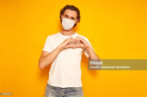 보호 의료 용 마스크를 쓴 젊은이 노란 배경에 고립 된 캐주얼 한 옷을 입은 청년은 사랑에 대한 감사의 표시로 가슴에 심장 모양으로 손을 접었습니다 사람 보안 건강의 개념