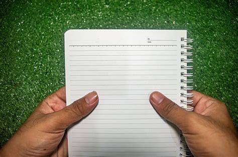 한 사람이 푸른 잔디 배경에 줄이 그어진 페이지가 있는 열린 나선형 노트북을 들고 있습니다 노트북 상단에는 아니요와 날짜를 입력할 수 있는 공간이 있어 일기를 쓰거나 메모를