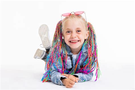 화려한 머리띠와 분홍색 선글라스를 끼고 웃고 있는 행복한 어린 아이가 스튜디오에서 하얀 배경에 누워 재미있는 머리띠와 분홍색 선글라스를 착용한 데님 의상을 입고 웃고 있다