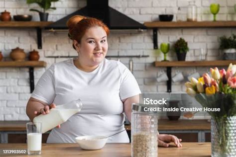 부엌에서 우유와 귀리 조각으로 만든 건강한 아침 식사를 요리하는 쾌활한 백인 여성 플러스 크기 통통 한 여자 다이어트 에 대 한
