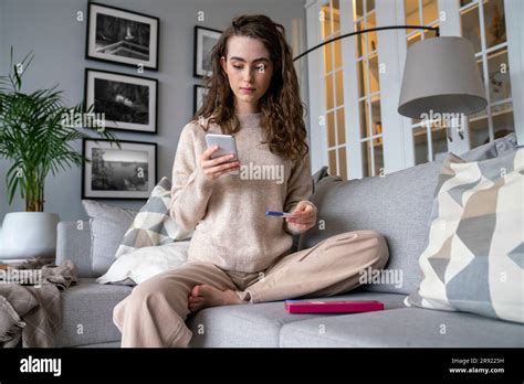 Young Woman Using Smart Phone Holding Pregnancy Testing Kit At Home