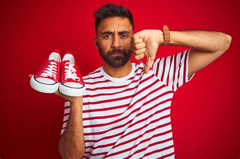 젊은 아랍 인도 히스패닉 남자 들고 스포츠 캐주얼 운동화 위에 고립 된 빨간 배경 화가 얼굴 엄지 손가락으로 싫어하는 표시 부정적인 기호 거부 개념 개념에 대한 스톡 사진