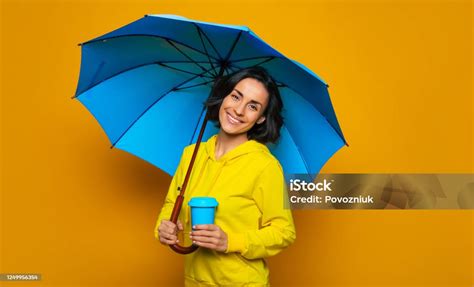 비오는 날을 즐기고 노란 후드를 입고 왼손에 파란색 열잔을 들고 우산 아래 얼굴에 넓은 미소를 지은 달콤한 소녀의 반 길이 사진