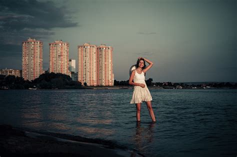 고층 건물의 배경에 젊은 여자 흰색 여름 드레스와 밀짚 모자를 입고 아름다운 행복한 금발이 해변의 물 위에 서서 미소짓고 있습니다 강에 대한 스톡 사진 및 기타 이미지