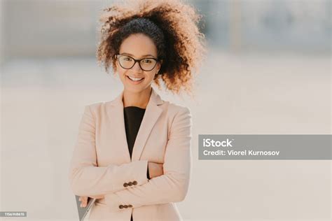 곱슬 한 아프로 머리와 자신감 쾌활한 여성 기업가 팔을 접힌 유지 현대 태블릿 컴퓨터를 사용 긍정적으로 미소 비즈니스 파트너 야외 대기 배경에 흐리게 도시 설정 고름에 대한