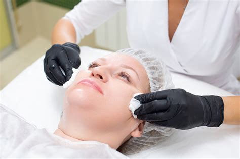 미용사 사무실에서 소파에 있는 의료용 모자에 누워 있는 한 젊은 여성의 초상화가 미용 시술 중 환자의 얼굴에서 면 패드로 과도한 화장품을 제거합니다 2명에 대한 스톡 사진