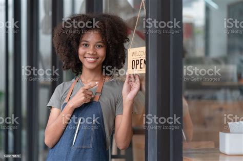 카페테리아 문 입구에 서 있는 아프리카계 미국인 사업가의 초상화 열린 간판이 있는 유리문 근처에 파란색 앞치마를 두르고 카메라를 바라보는 쾌활한 젊은 웨이트리스 20 24