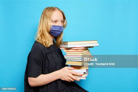 가면을 쓴 백인 학생 소녀가 커다란 산의 책과 함께 앉아 있다 코로나바이러스 중 격리 및 온라인 학습 개념 밝은 파란색 배경에