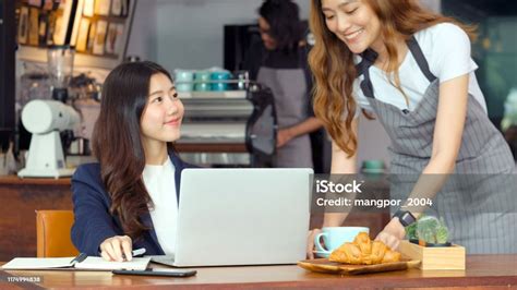 젊은 아시아 여자 바리스타 제공 A 커피 컵 와 미소 에 사업가 고객 동안 그녀 작업 에 노트북 컴퓨터 에 카페 커피 숍 배경