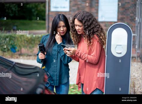 Woman Holding Electric Plug And Sharing Mobile Phone With Friend At Charging Station Stock Photo