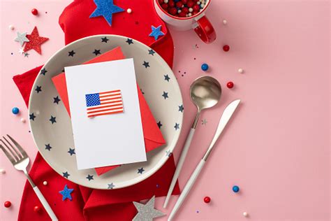 메모리얼 데이 파티 설정 접시 수저 컵 냅킨 미국 국기가 있는 엽서 봉투 반짝이는 별 스프링클 파스텔 핑크 배경에 색종이 조각 등 애국적인 장식으로 장식된 테이블의 평면도