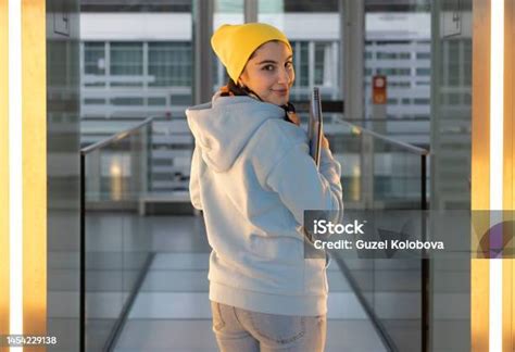 파란색 스웨트셔츠와 노란색 모자를 쓴 웃는 여학생이 유리 복도를 따라 걷는다 캠퍼스에 대한 스톡 사진 및 기타 이미지 캠퍼스