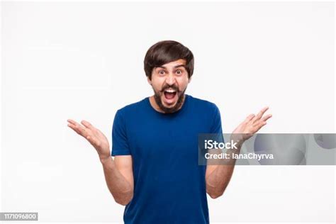 수염과 가장 큰 수염이있는 파란색 셔츠를 입은 브루넷 남자가 흰색 배경 위에 고립 된 서 있는 것을 보고 어깨를 으쓱하고 있습니다 감정에 대한 스톡 사진 및 기타 이미지