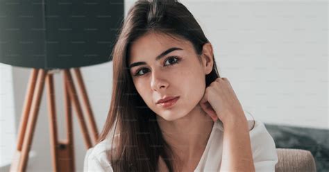 매력적인 여자가 회색 램프 근처의 의자에 앉아 있다 하얀 벽과 창문에서 나오는 일광이 있는 방에 있는 젊은 갈색 머리 사진