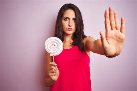 진지하고 자신감있는 표정 방어 제스처로 정지 표지판을하는 오픈 손으로 분홍색 고립 된 배경을 통해 달콤한 사탕을 먹는 젊은 아름다운 여자 경계 표지에 대한 스톡 사진 및