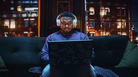 아늑한 세련된 로프트 아파트의 노트북 컴퓨터에서 집에서 일하는 헤드폰의 젊은 잘 생긴 흑인 창조적 인 남성 미소 소셜 미디어 확인 인터넷 검색 큰 창문으로 평평합니다