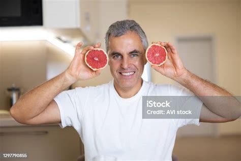 더 많은 비타민 잘 생긴 남자는 얼굴 근처에 자몽의 절반을 잘라 들고 이빨로 미소와 카메라를보고 건강한 식품 개념 배경에 아늑한 현대적인 부엌 50 54세에 대한 스톡 사진