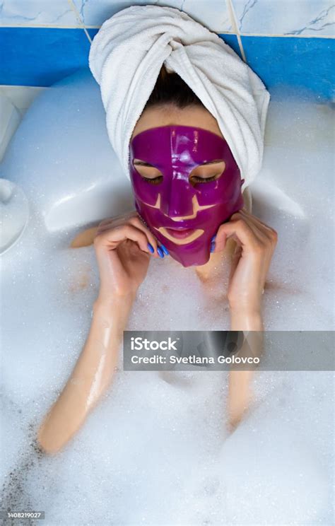 머리에 수건을 씌운 어린 소녀가 욕실에 누워 있고 얼굴에 분홍색 얼굴 마스크가 있습니다 소녀는 욕실에 누워 있습니다 그녀의