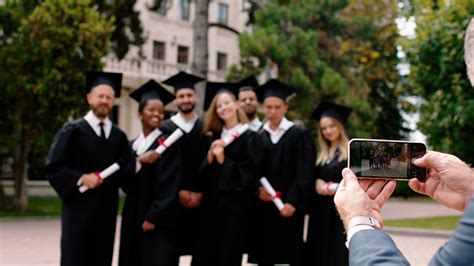 다인종 졸업생 그룹의 졸업식 날 대학 공원에서 스마트폰 앞에서 큰 미소를 지으며 포즈를 취하는 학생들 부모 중 한 명이 추억을 위해 사진을 찍는 노인 4k해상도에 대한 스톡