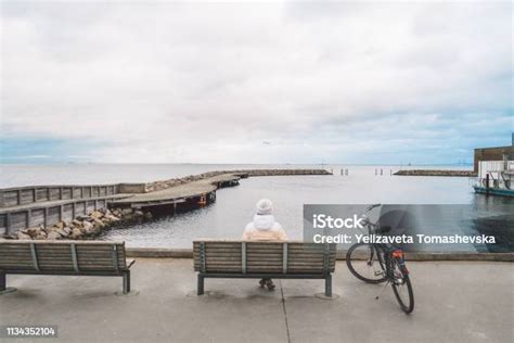 젊은 백인 여자는 흐린 날씨에 겨울에 코펜하겐 덴마크의 바닷가에 발트 해를 내려다 볼 수 있는 나무 벤치에 그녀의 뒤에 앉아