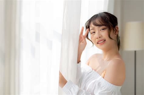 아침에 일출과 함께 일어나면서 창에 서있는 아름다운 젊은 아시아 여성의 초상화 신선함과 쾌활한 라이프 스타일과 휴식 개념에 만족하는 소녀 가정 생활에 대한 스톡 사진 및