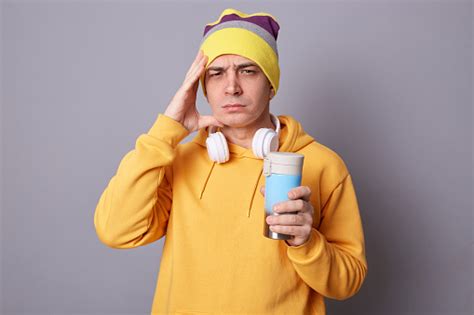 노란색 후드티와 비니 모자를 쓴 힙스터 남자의 실내 샷 관자놀이에 손을 대고 얼굴을 찡그리고 두통을 앓고 에너지가 필요하고 보온병에서 커피를 마시고 회색 벽에 서 있습니다