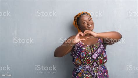 심장 기호와 손으로 모양을 보여주는 사랑에 웃 고 오렌지 머리 띠를 입고 회색 그런 지 벽에 젊은 아프리카계 미국인 여자