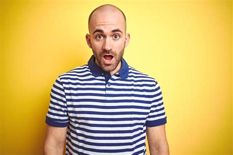 노란색 고립 된 배경에 캐주얼 스트라이프 블루 티셔츠를 입고 수염을 가진 젊은 대머리 남자는 놀라움의 표정 두려움과 흥분 된